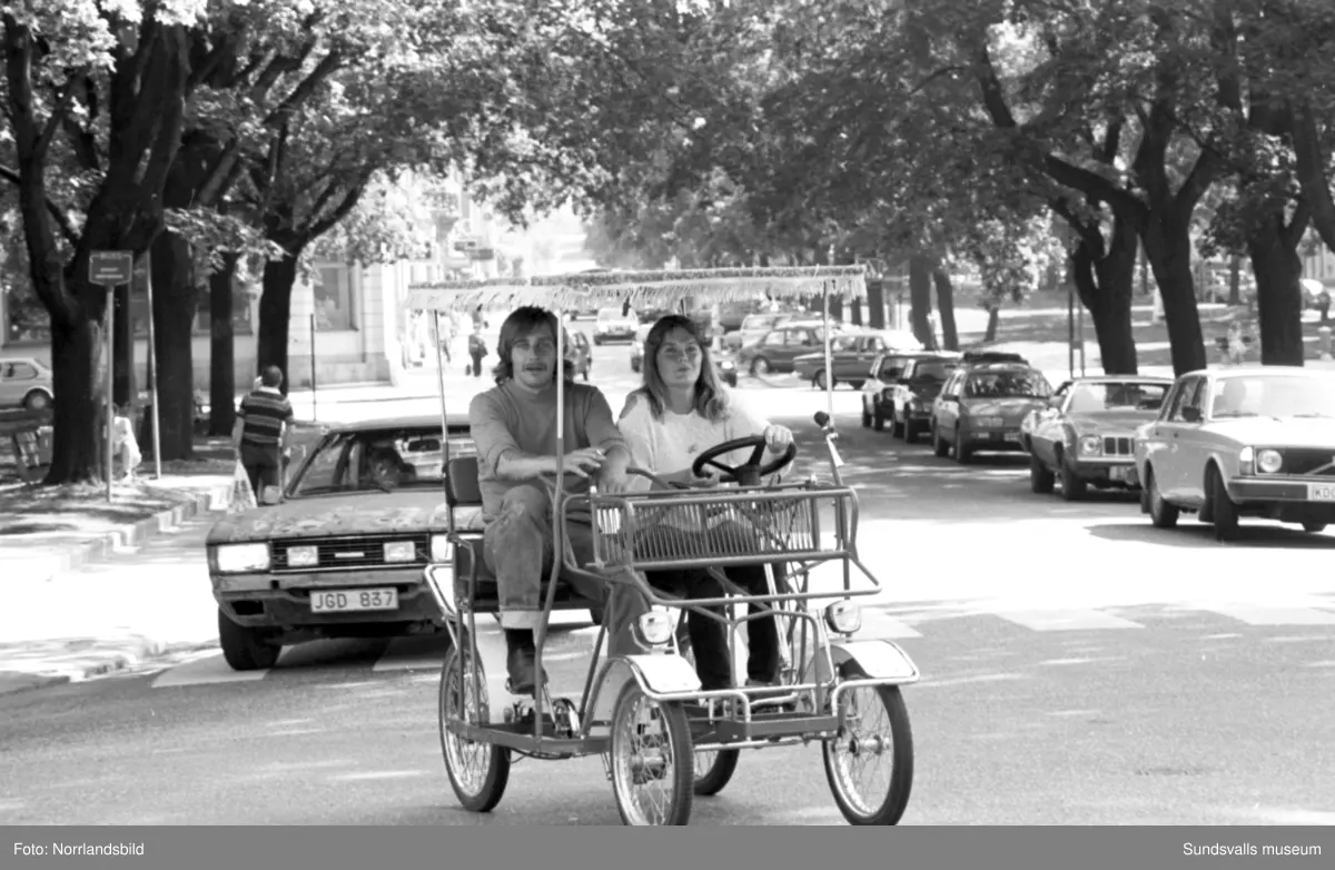 "Cykelbil." Ett ungt par framför en slags fyrhjulig cykel med tak och ...