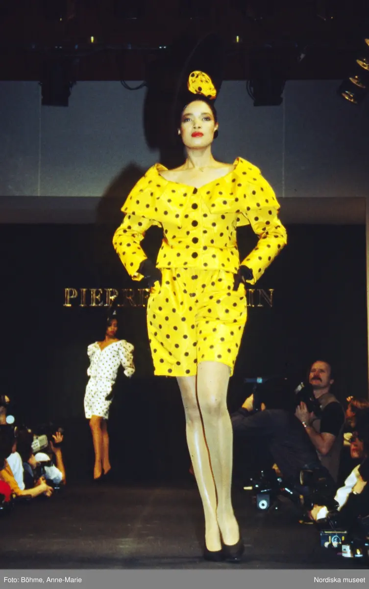 Modevisning. Modell i gul dräkt med svarta prickar och matchande hatt ...