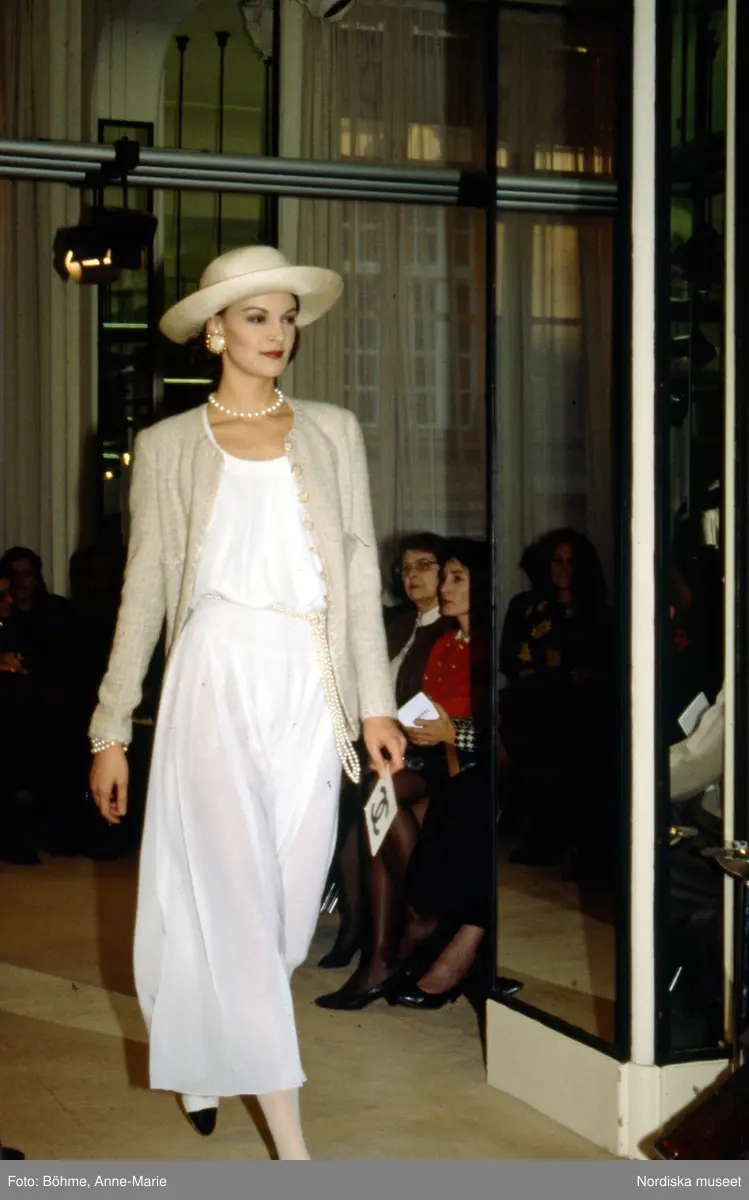 Modevisning. Modell i vit klänning med knutet band av pärlor i midjan ...