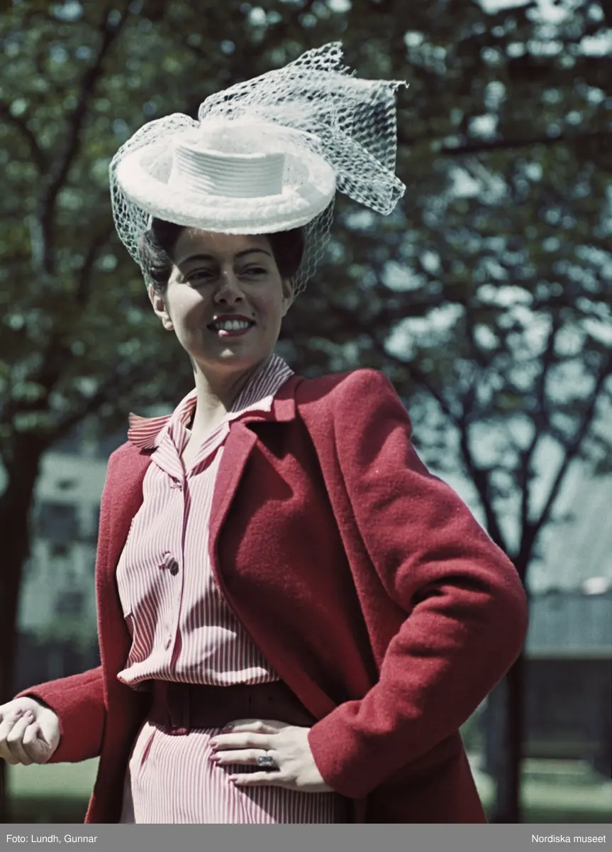 Kvinna i rödrandig klänning med skärp, vit hatt med flor och röd kavaj ...