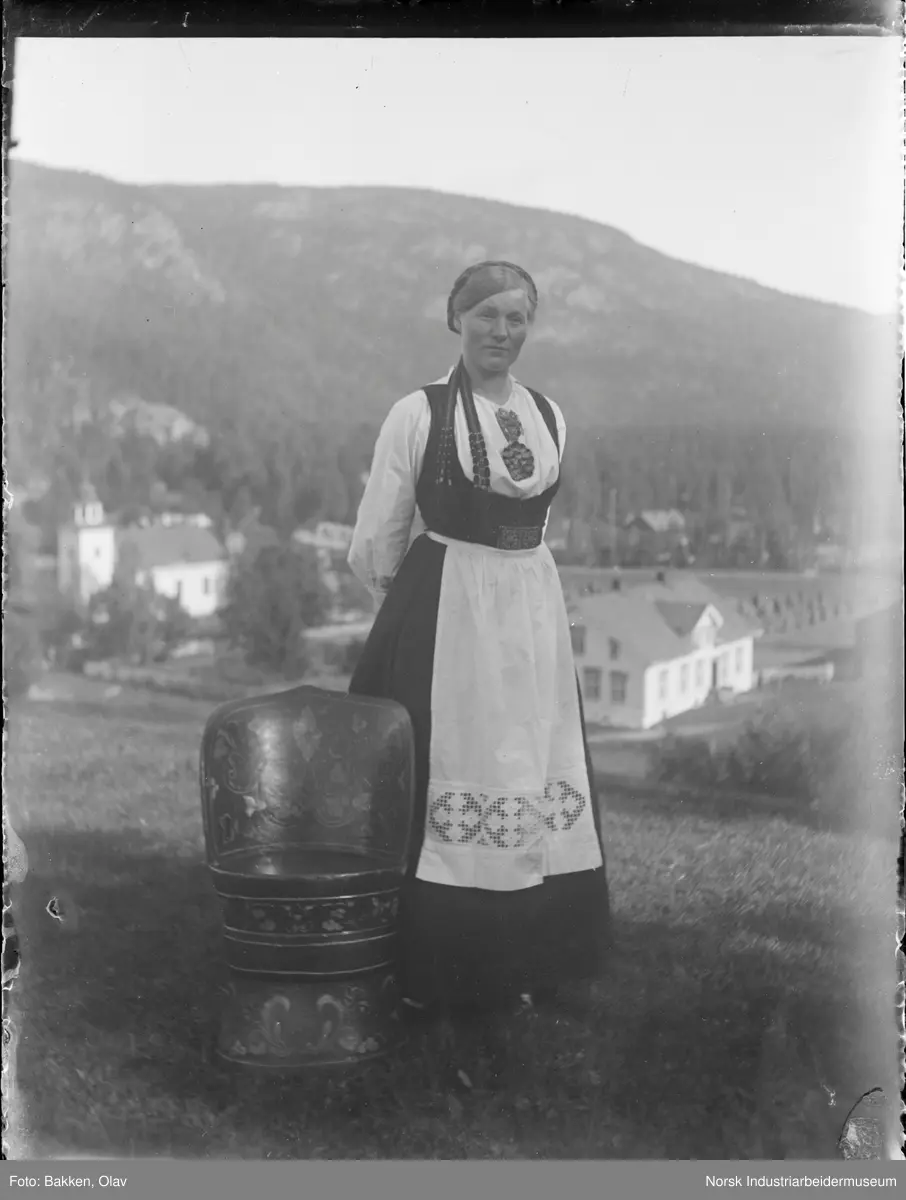 Portrett. Kvinne stående i bunad ved siden av kubbestol utendørs ...