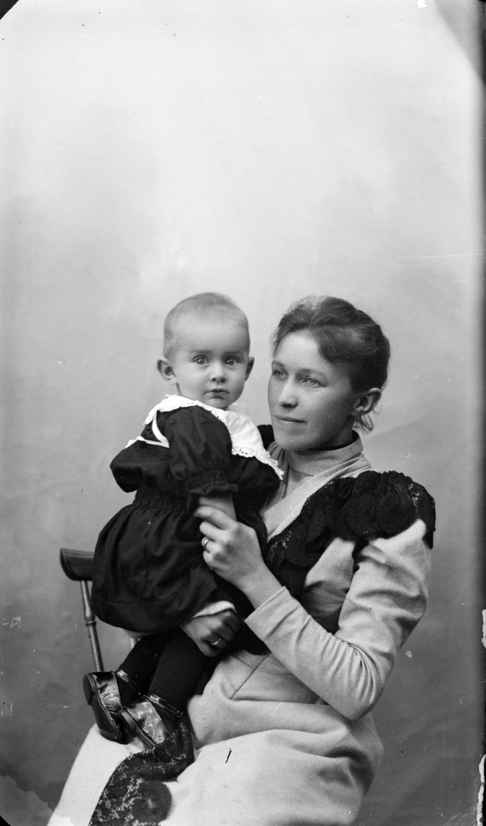 Mor med barn på armen, antatt 1 år, halvfoto. Barnet har mørk kjole med ...