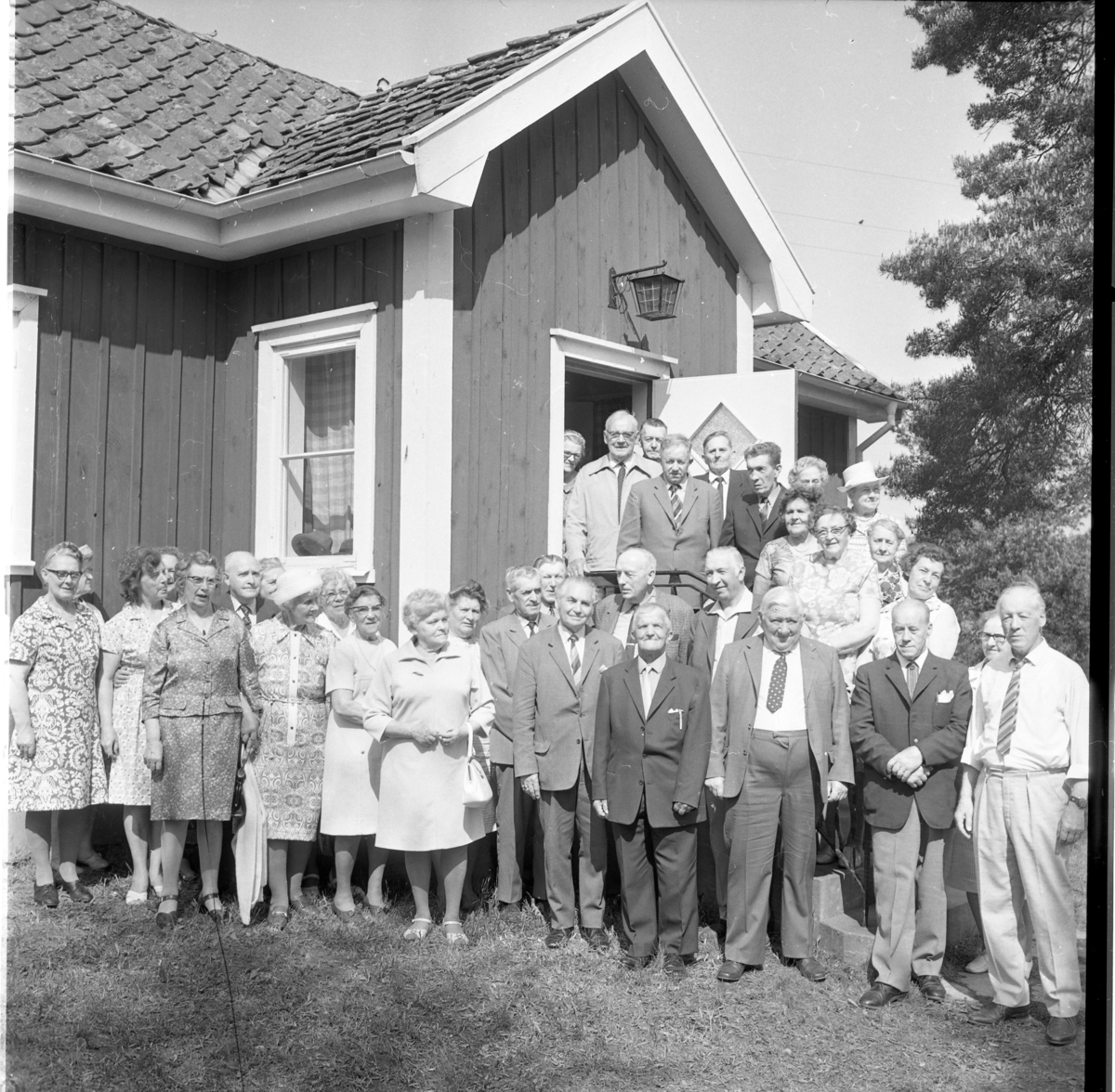 En stor grupp äldre kvinnor och män framför en rödfärgad stuga med vita ...