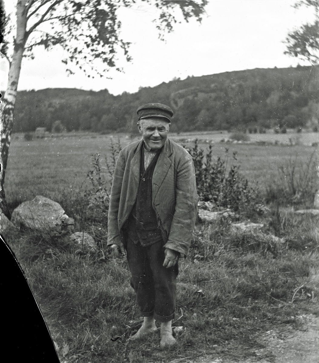 En man står barfota utomhus i gräset med åkerlandskap och berg i ...
