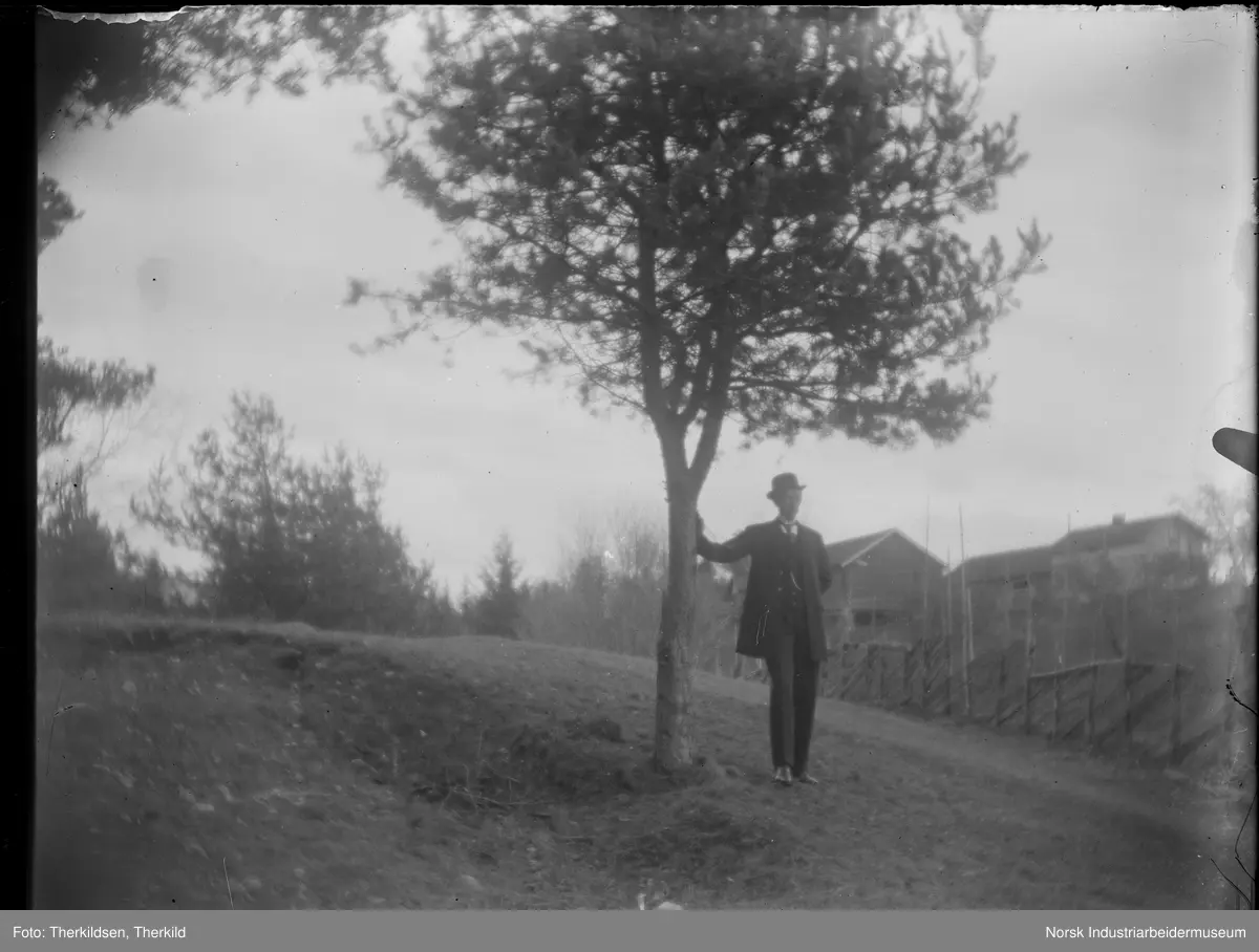 Mann lener seg mot en trestamme på et jorde. Gårdsbygninger i ...