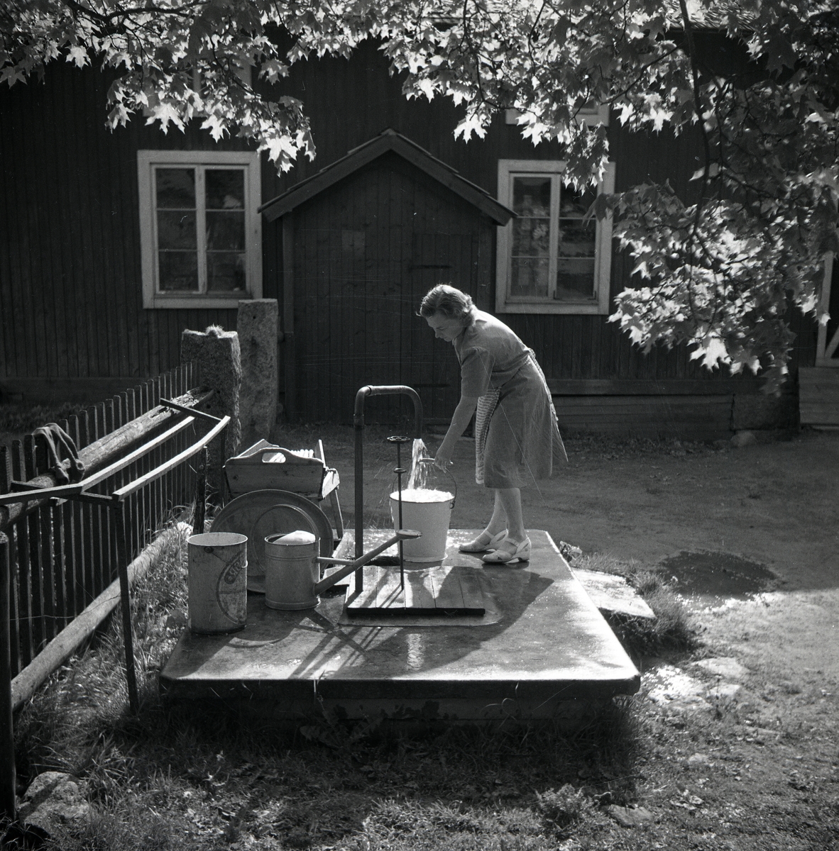 Adéle Mickelsson står och fyller vatten i en hink vid en gårdsbrunn ...