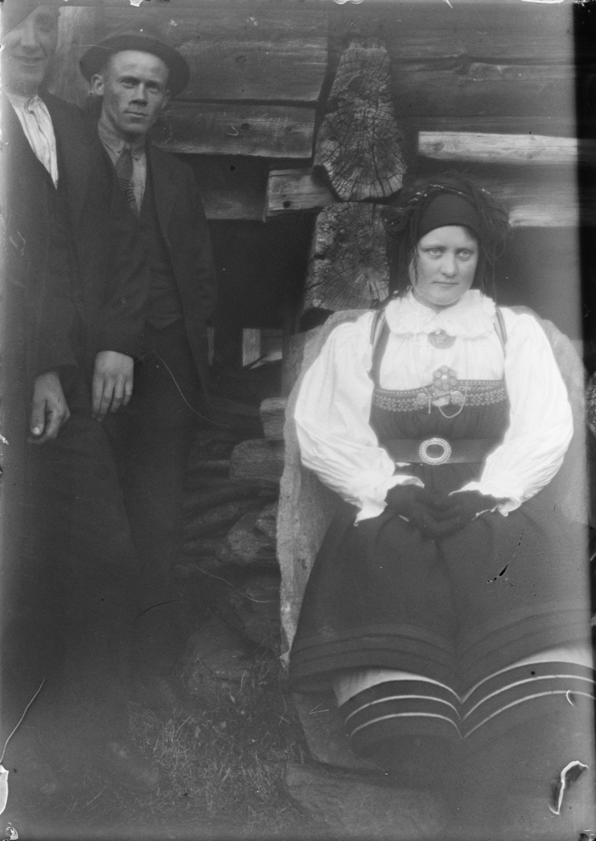 Kvinne i setesdalsbunad og to unge menn bak. Trolig Voss folkehøgskule ...