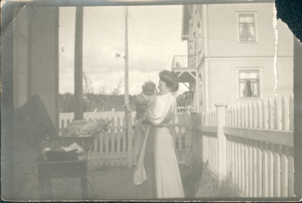 Gruppe. Fru Rasmus Dahl, holder en baby. I bakgrunnen gjerdet til ...